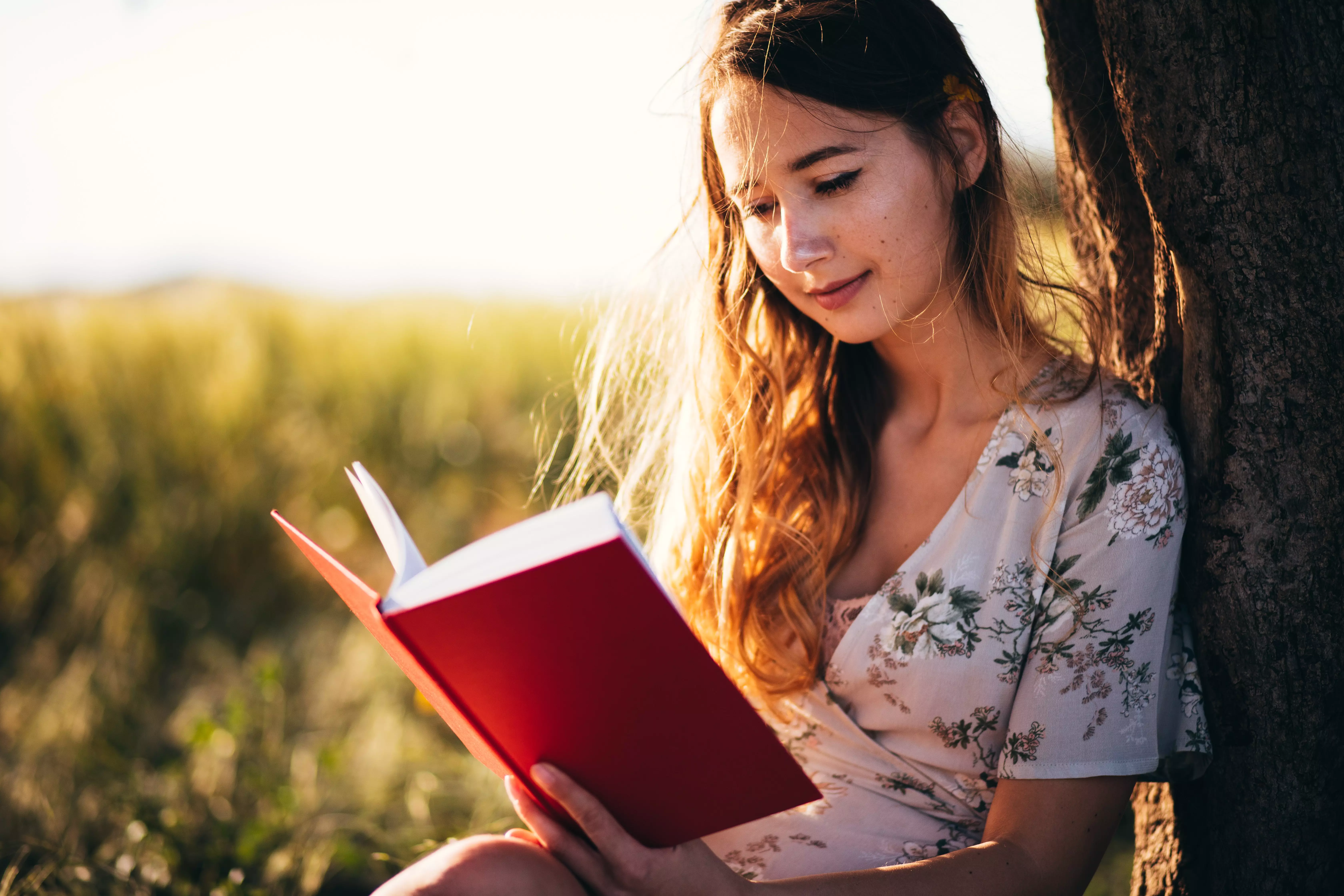 Çiçəkli paltar geyinmiş qadın ağacın yanında oturur və gün batarkən kitab oxuyur.