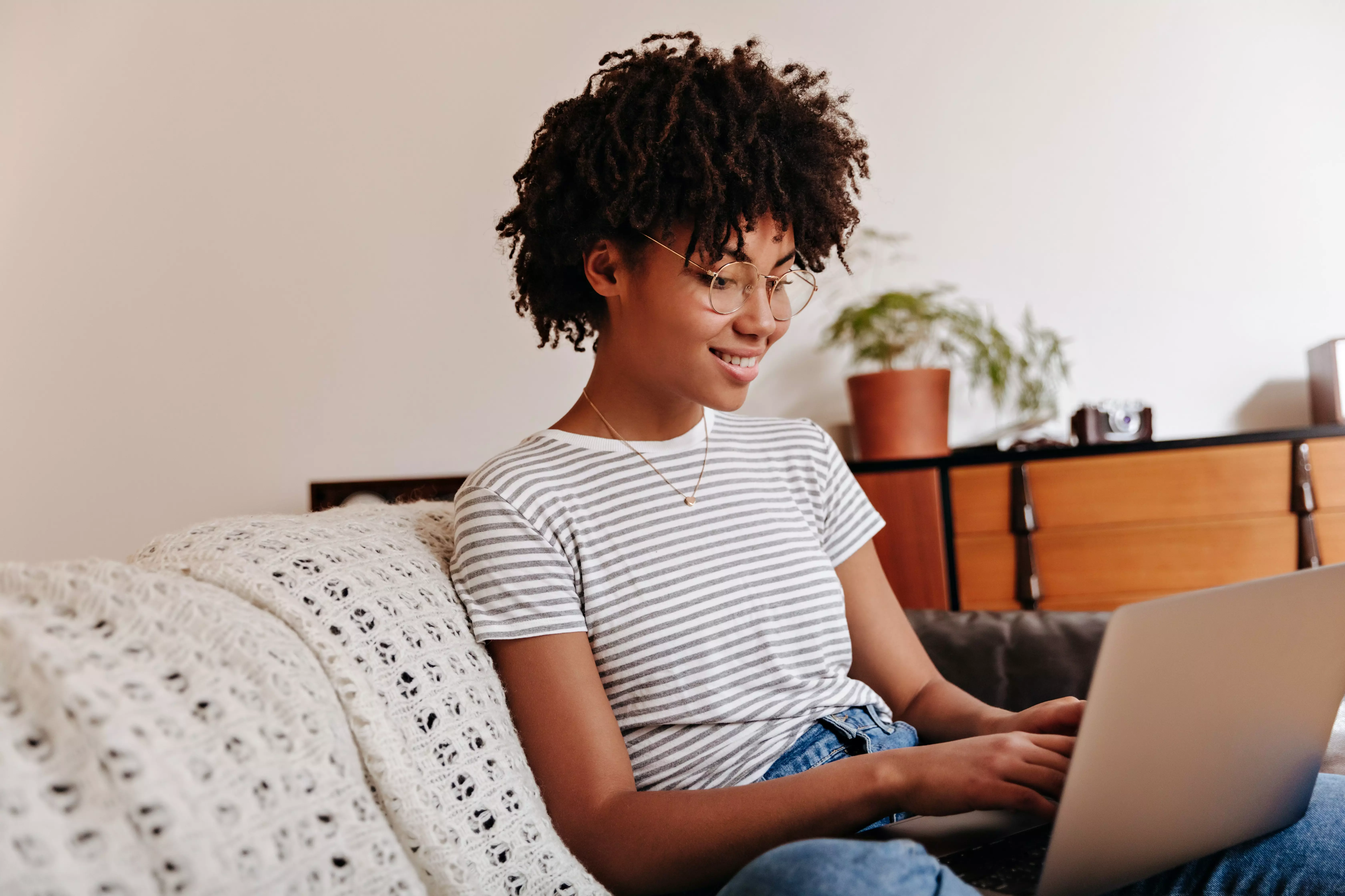 Eng Fra, déi e gesträifte Hiem an Jeans unhuet, sëtzt op hirer Canapé wärend se vun doheem op hirem Laptop an ...