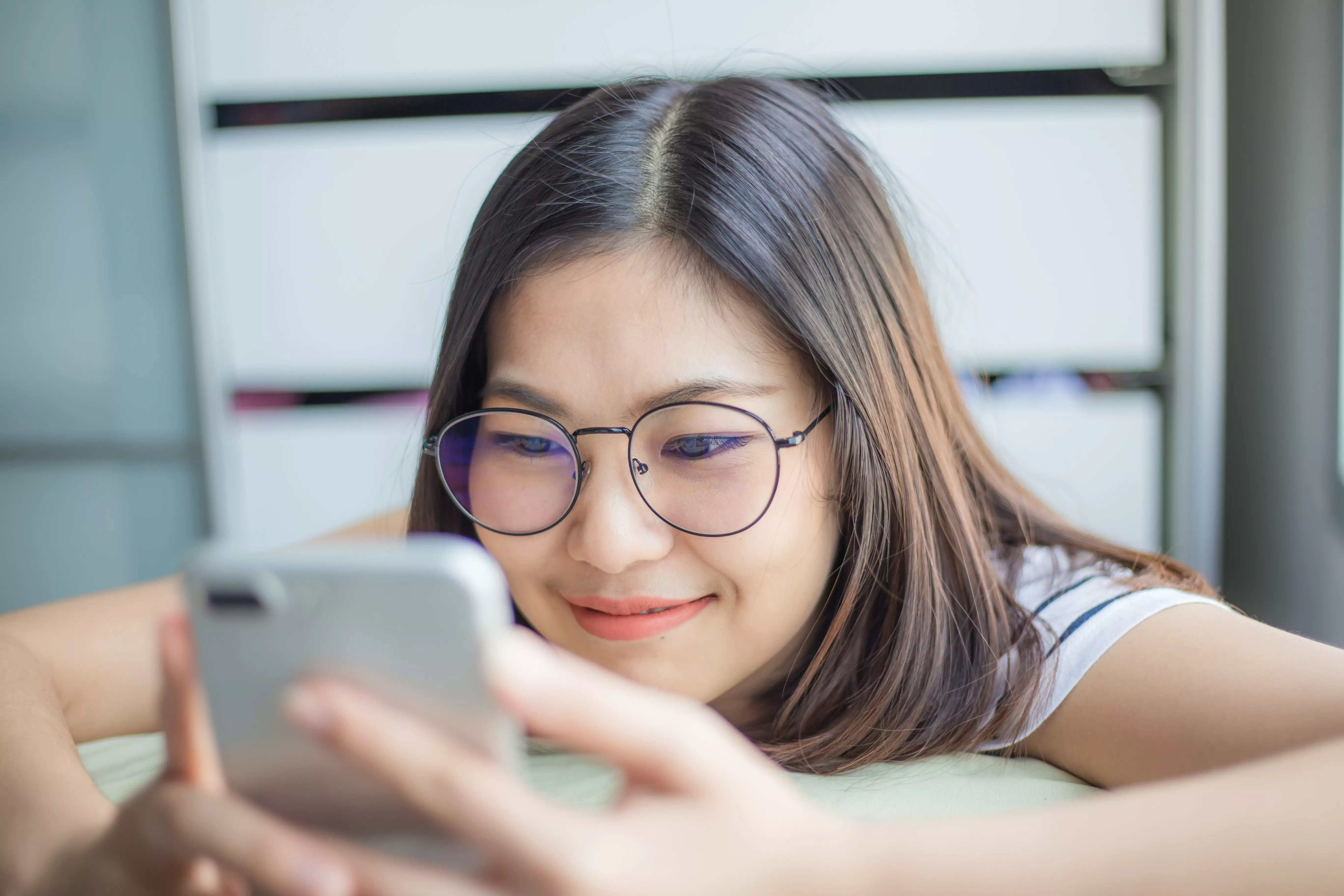 Fröhliche junge Brillen lächelnde asiatische Frauen mit Smartphone auf gemütlichem Bett SMS-Nachrichten